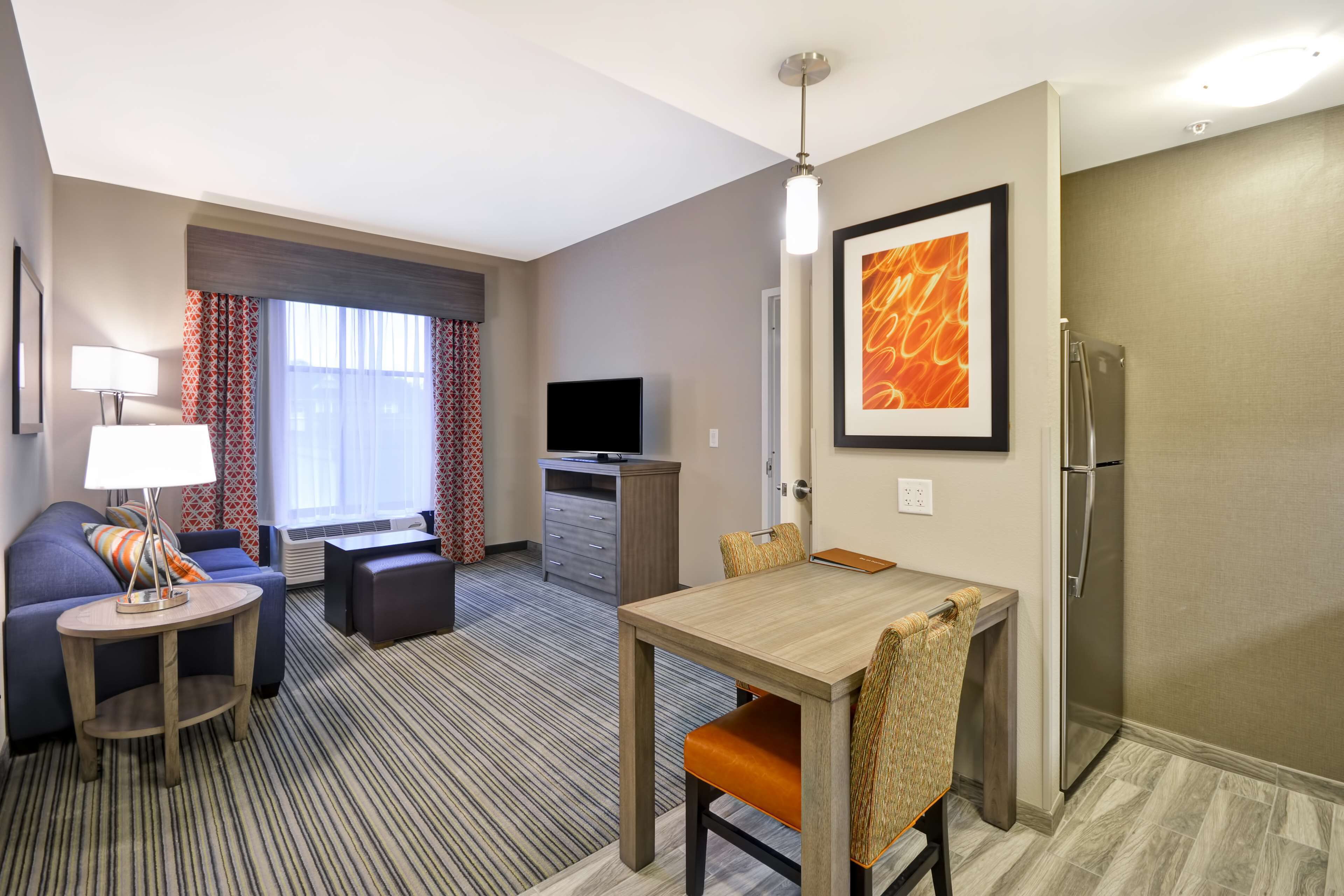 Quarto de hotel com sofá, mesinha com luminária, televisão, mesa de jantar com cadeiras e geladeira.