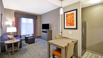 Quarto de hotel com sofá, mesinha com luminária, televisão, mesa de jantar com cadeiras e geladeira.