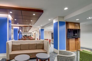 Um hall de hotel moderno com área de estar, espaço para refeições e uma TV fixada em um painel de madeira.