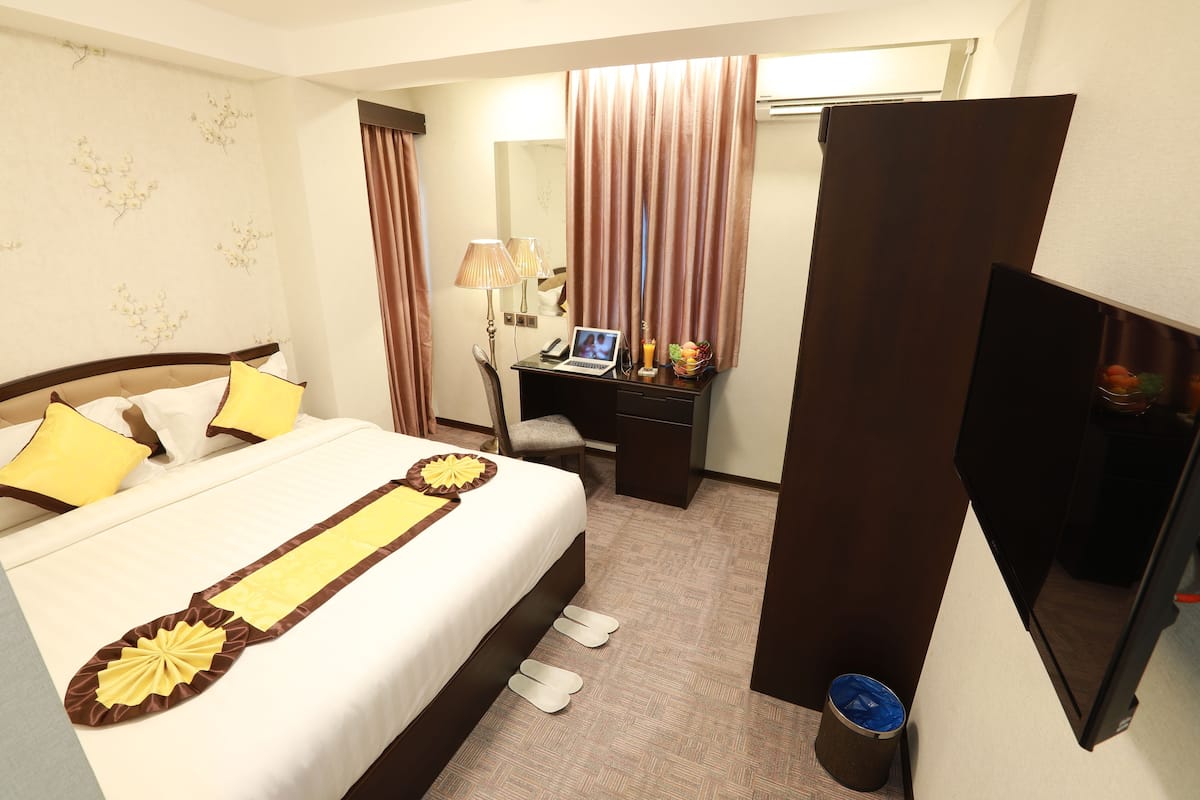 Quarto de hotel com cama, escrivaninha, cadeira, televisão e lixeira.