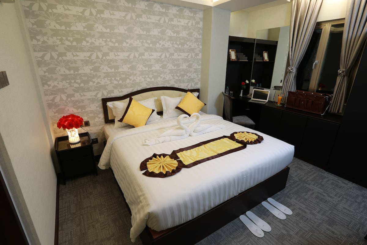 Uma cama bem arrumada com lençóis brancos e almofadas decorativas, uma mesinha de cabeceira com abajur e um vaso de flores, e um espelho grande na parede.