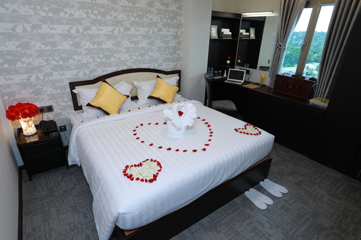 Cama bem arrumada com roupa de cama branca, arranjos florais em formato de coração e um centro de mesa em forma de cisne. Há uma escrivaninha com um laptop e um espelho, além de uma janela com cortinas.