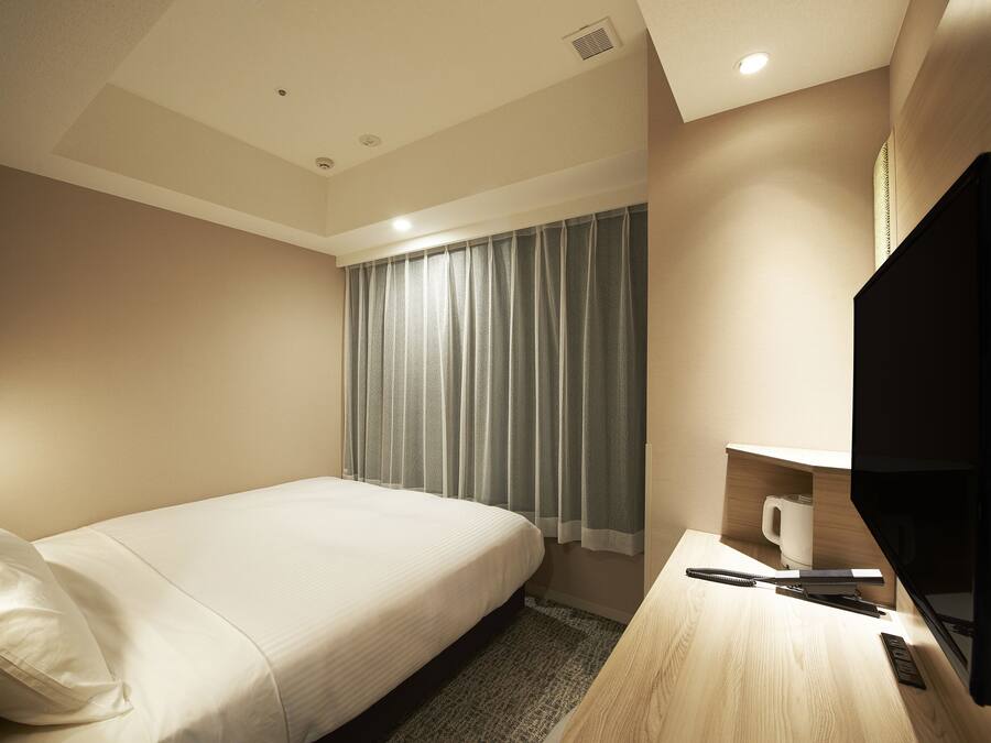 Quarto de hotel com uma cama de solteiro, escrivaninha com televisão, cafeteira e controle remoto.
