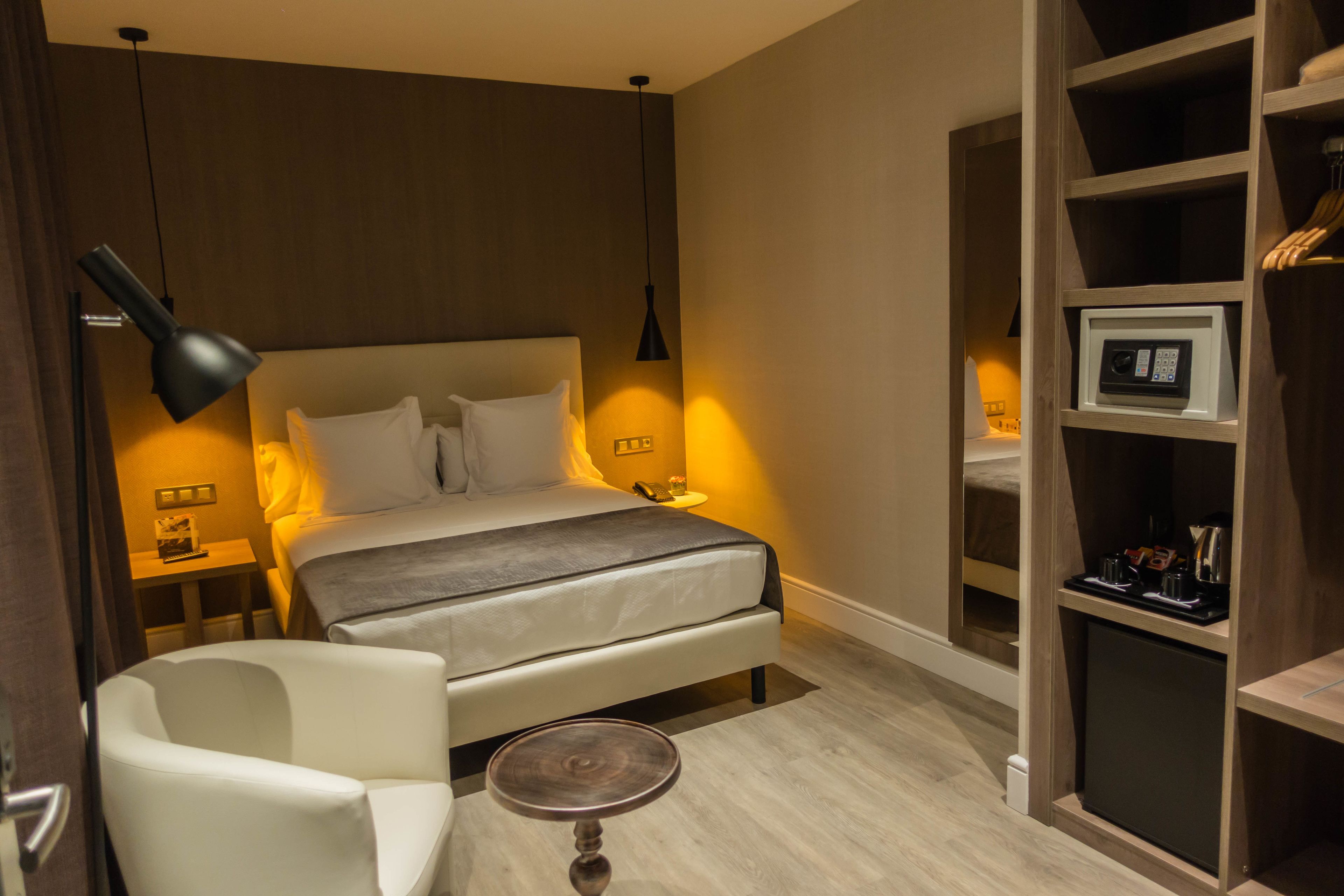 Quarto de hotel moderno com uma cama grande, abajures, uma mesa redonda pequena e um guarda-roupa com prateleiras.