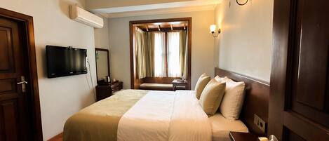 Quarto de hotel com cama, televisão, escrivaninha e janela com cortinas.