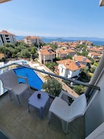 Uma varanda com piscina, cadeiras de descanso brancas e vista para uma área residencial com casas de telhado vermelho e um corpo d'água ao longe.