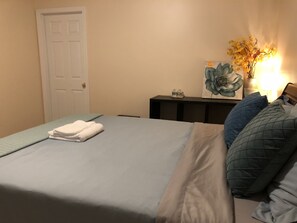 Cama bem arrumada com uma toalha branca dobrada, mesa de cabeceira com abajur e uma obra de arte na parede.