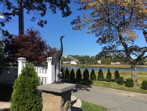 Um parque com uma estátua de um pássaro sobre um pedestal, um lago e casas ao fundo.