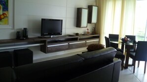 Uma sala de estar moderna com televisão de tela plana, móveis de madeira escura e um espaço de jantar com mesa e cadeiras.