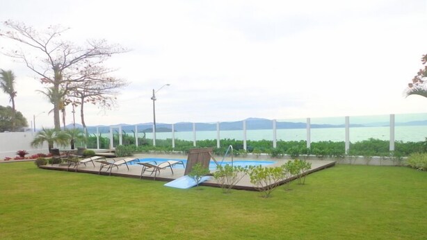 Uma área de piscina com vista para o mar, rodeada por um jardim bem cuidado e algumas árvores.