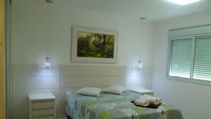 Um quarto com uma cama, duas mesas de cabeceira, um criado-mudo, uma janela com persianas e um quadro na parede.