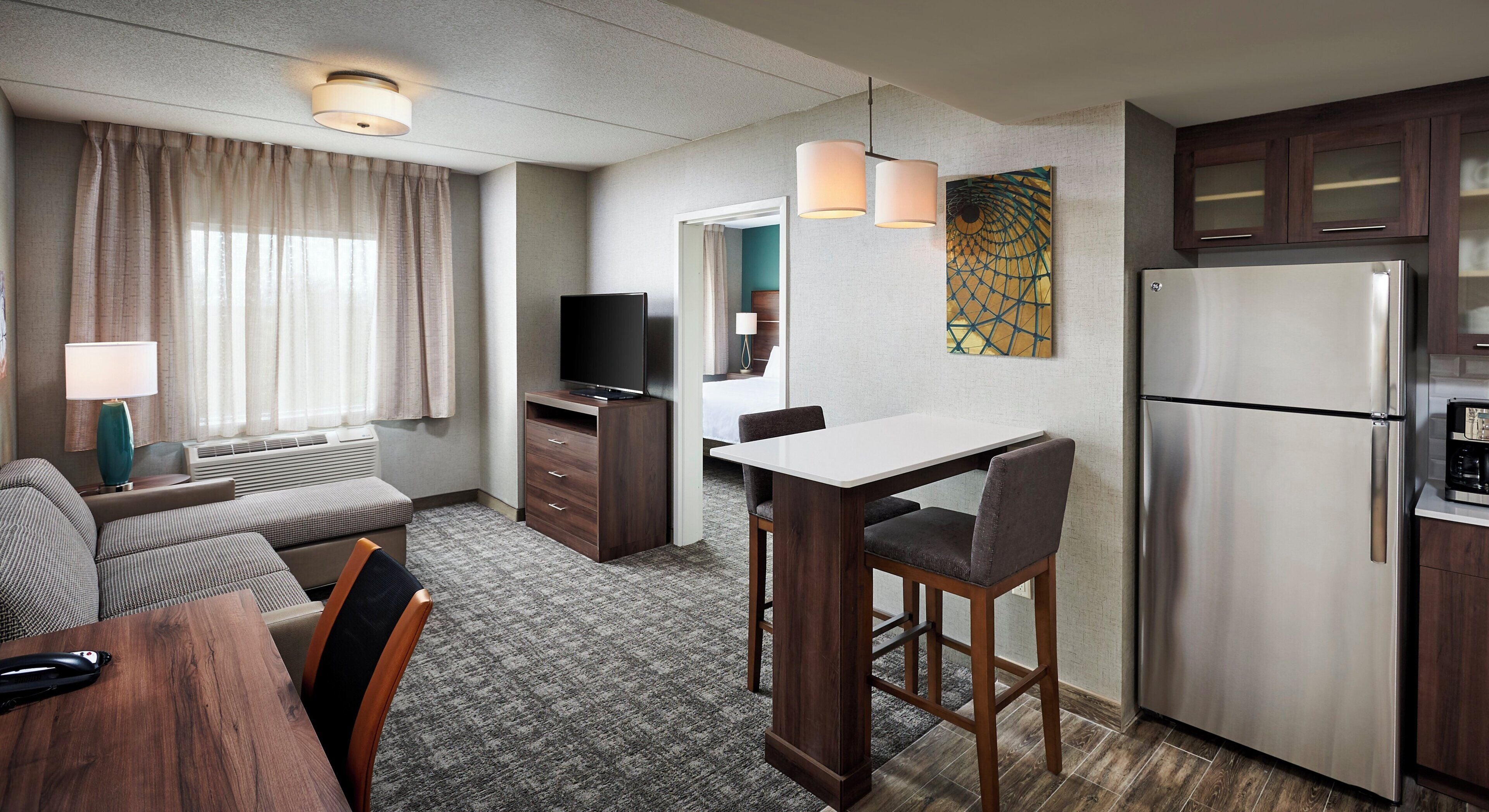 Quarto de hotel com cozinha compacta, área de refeições com mesa e cadeiras, sofá, televisão e cama ao fundo.