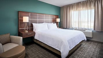 Quarto de hotel com uma cama grande, duas luminárias de cabeceira, uma mesinha de cabeceira, uma cadeira e uma janela com cortinas.
