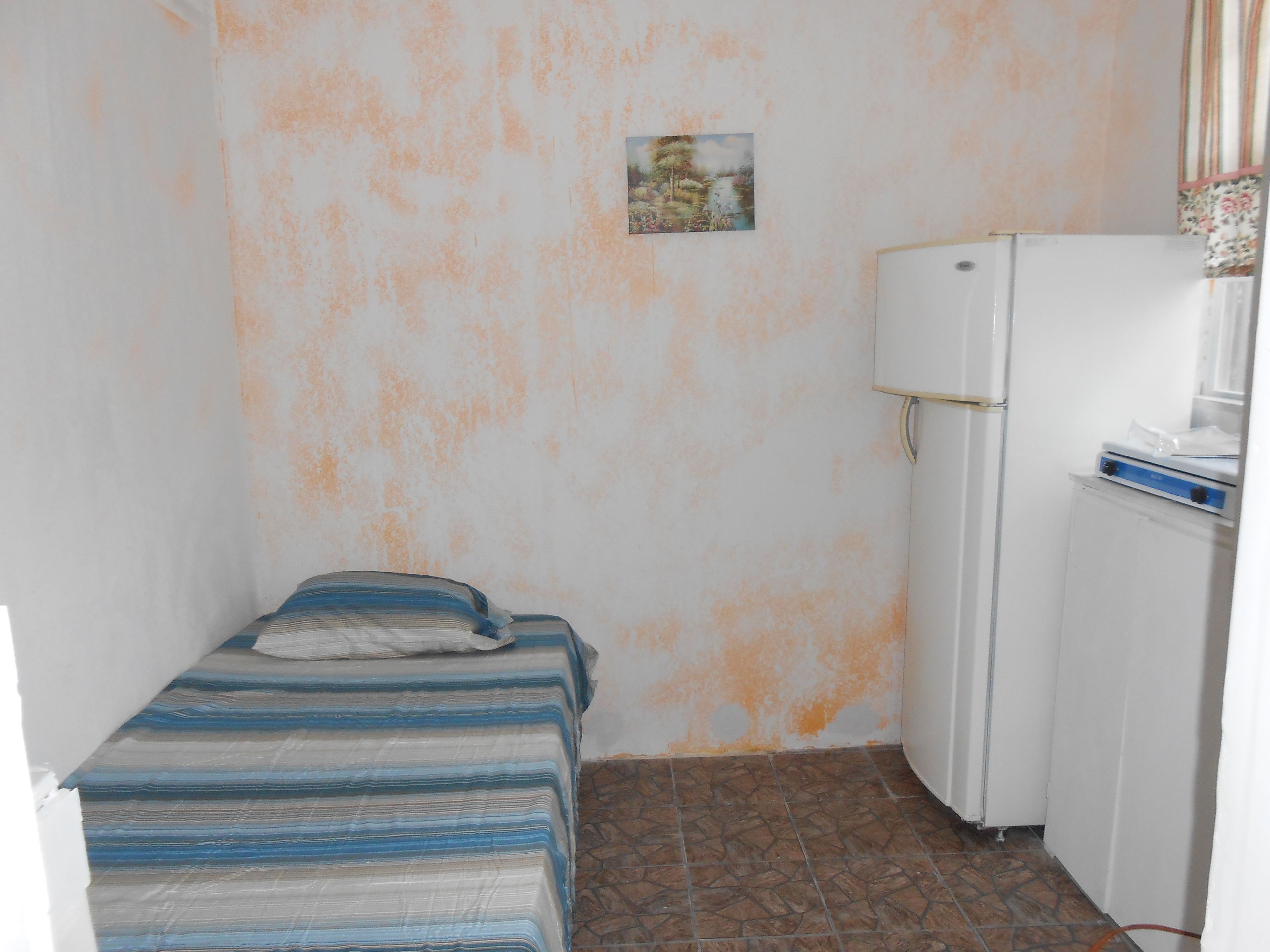 Um quarto pequeno e estreito com uma cama, um refrigerador e um pequeno quadro na parede.