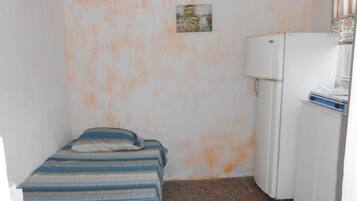 Um quarto pequeno e estreito com uma cama, um refrigerador e um pequeno quadro na parede.