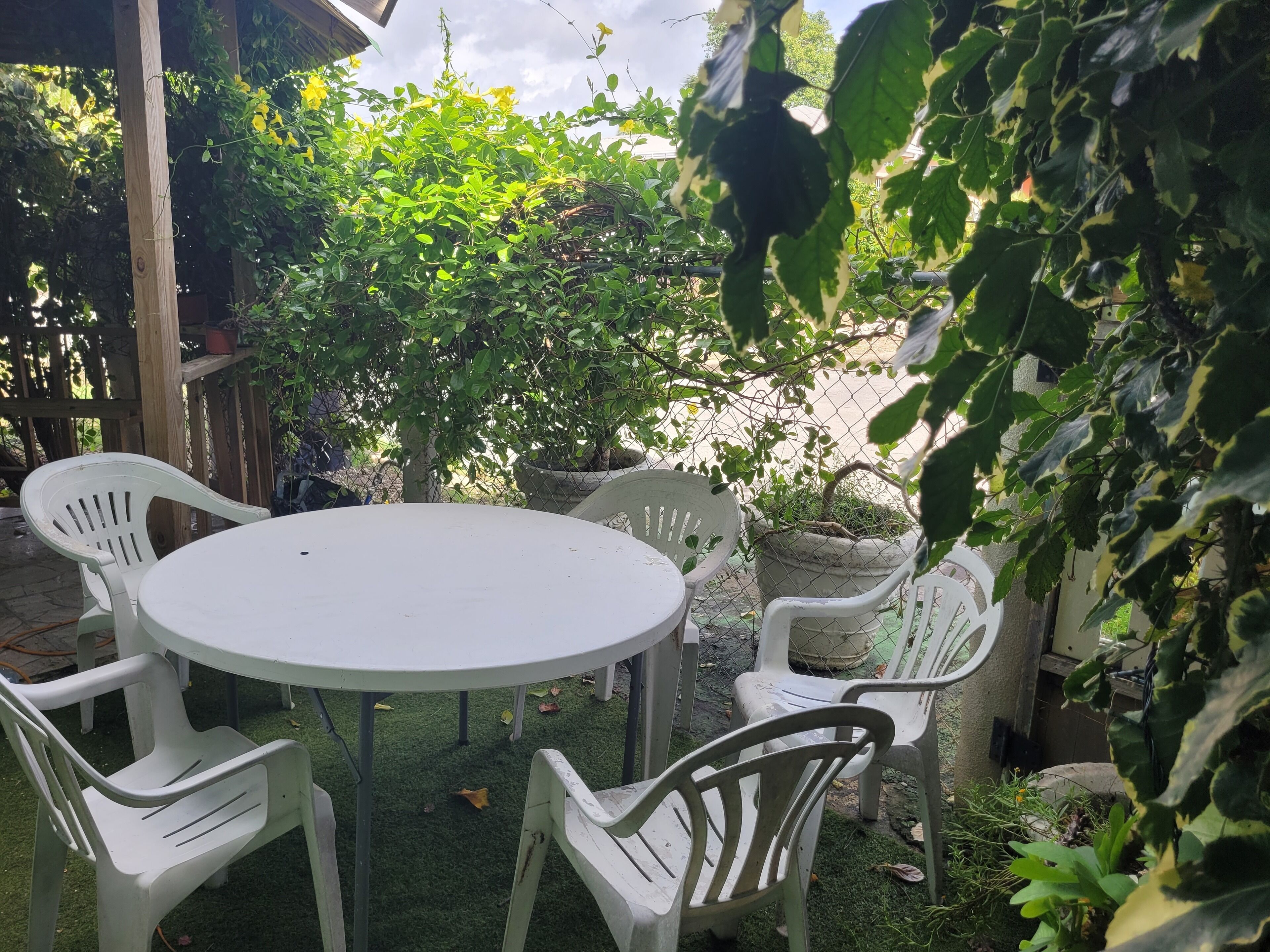 Uma mesa redonda com quatro cadeiras de plástico branco, cercada por vegetação e uma cerca de madeira.