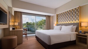 Quarto de hotel moderno com uma cama grande, uma escrivaninha, uma cadeira, televisão e varanda com vista para o verde.