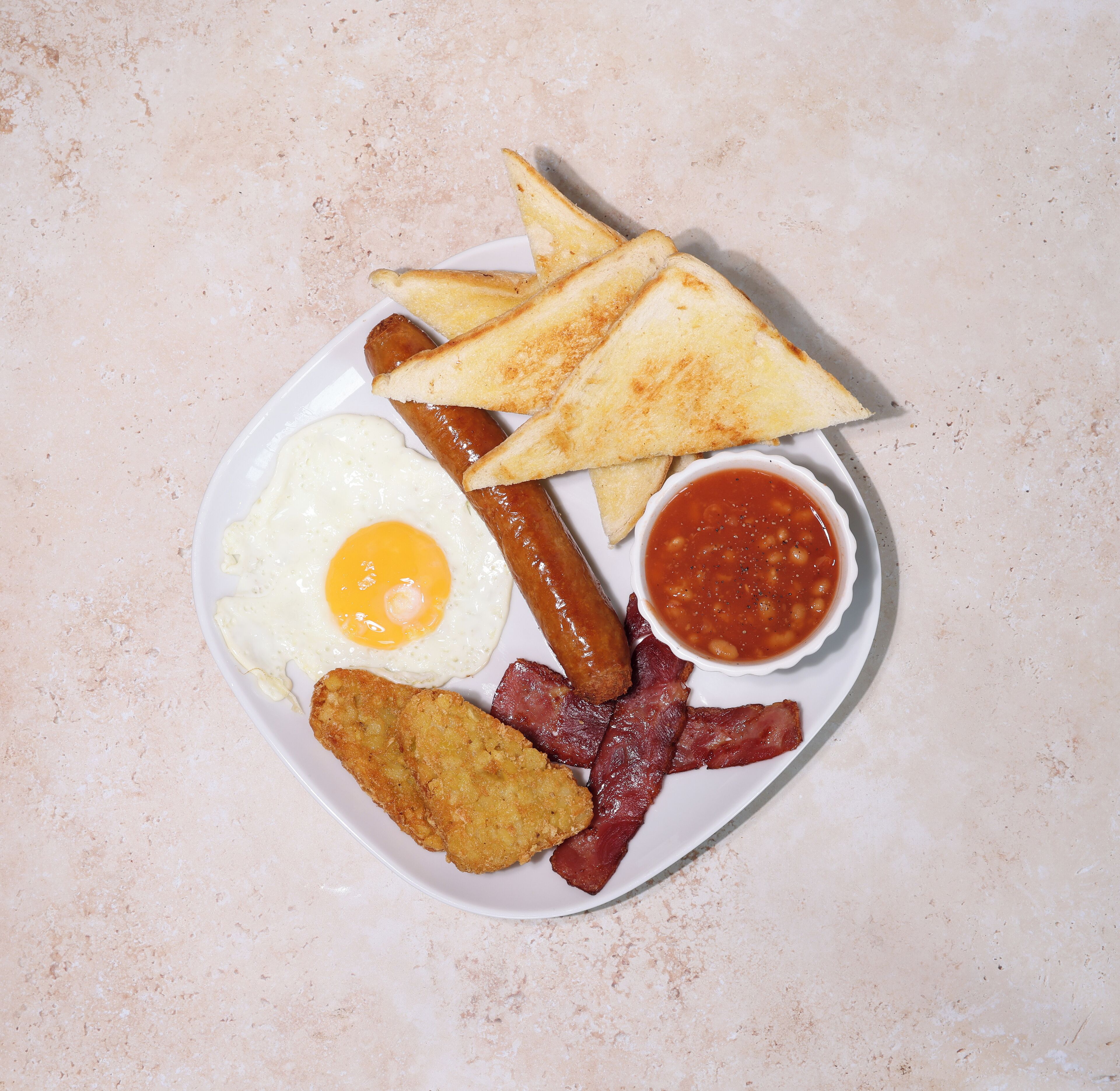 Um prato de comida de café da manhã com torradas, salsichas, bacon, um ovo e uma tigela de feijão.