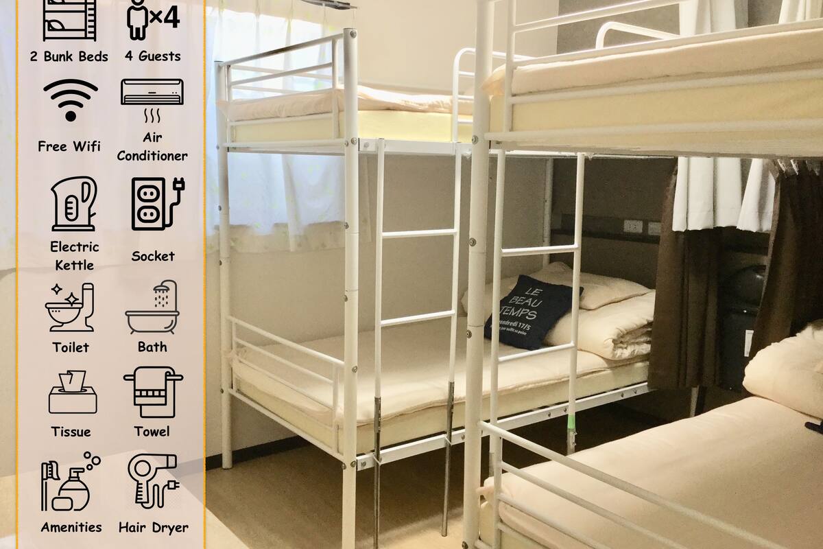 Beliches com estrutura metálica branca, uma janela com cortinas finas e uma toalha sobre uma das camas.