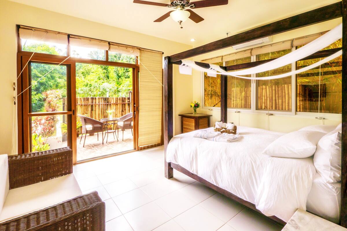 Quarto com uma cama grande, um ventilador de teto e uma porta de vidro deslizante que dá para uma área externa com mesa e cadeiras.