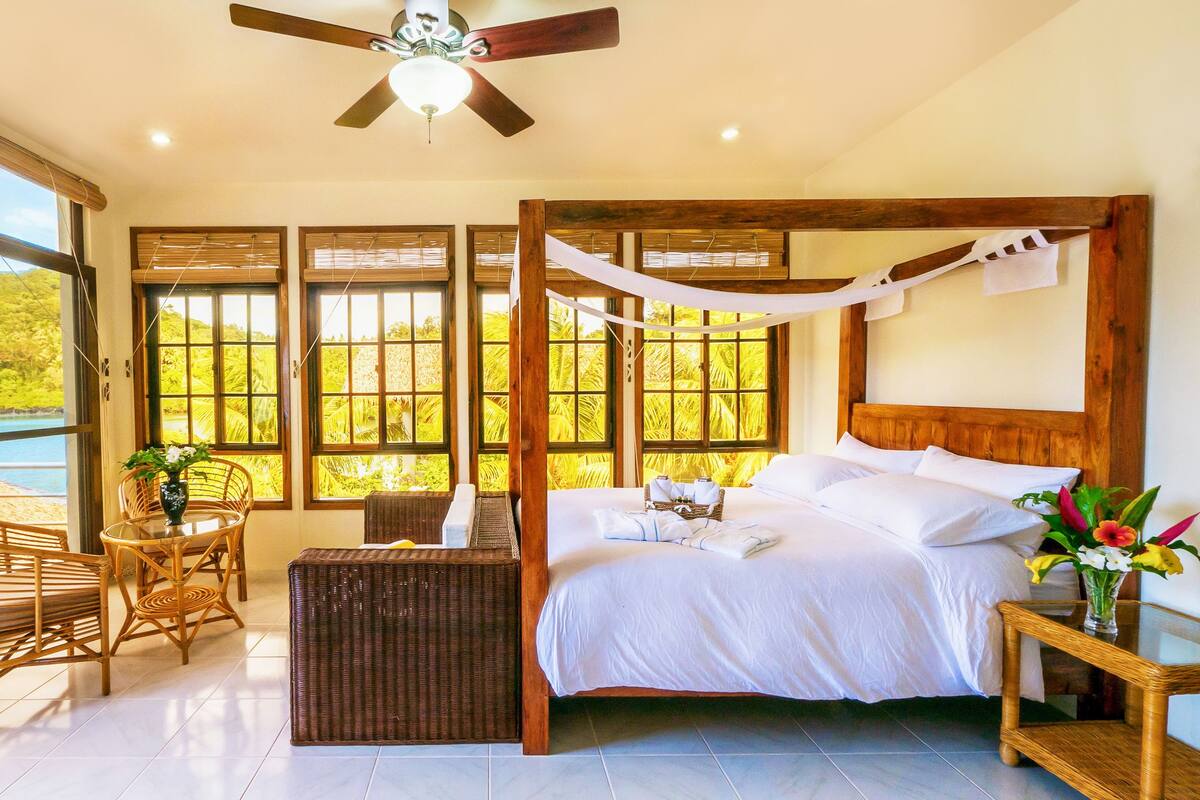 Quarto com uma cama grande, ventilador de teto e vista para uma paisagem tropical através de amplas janelas.