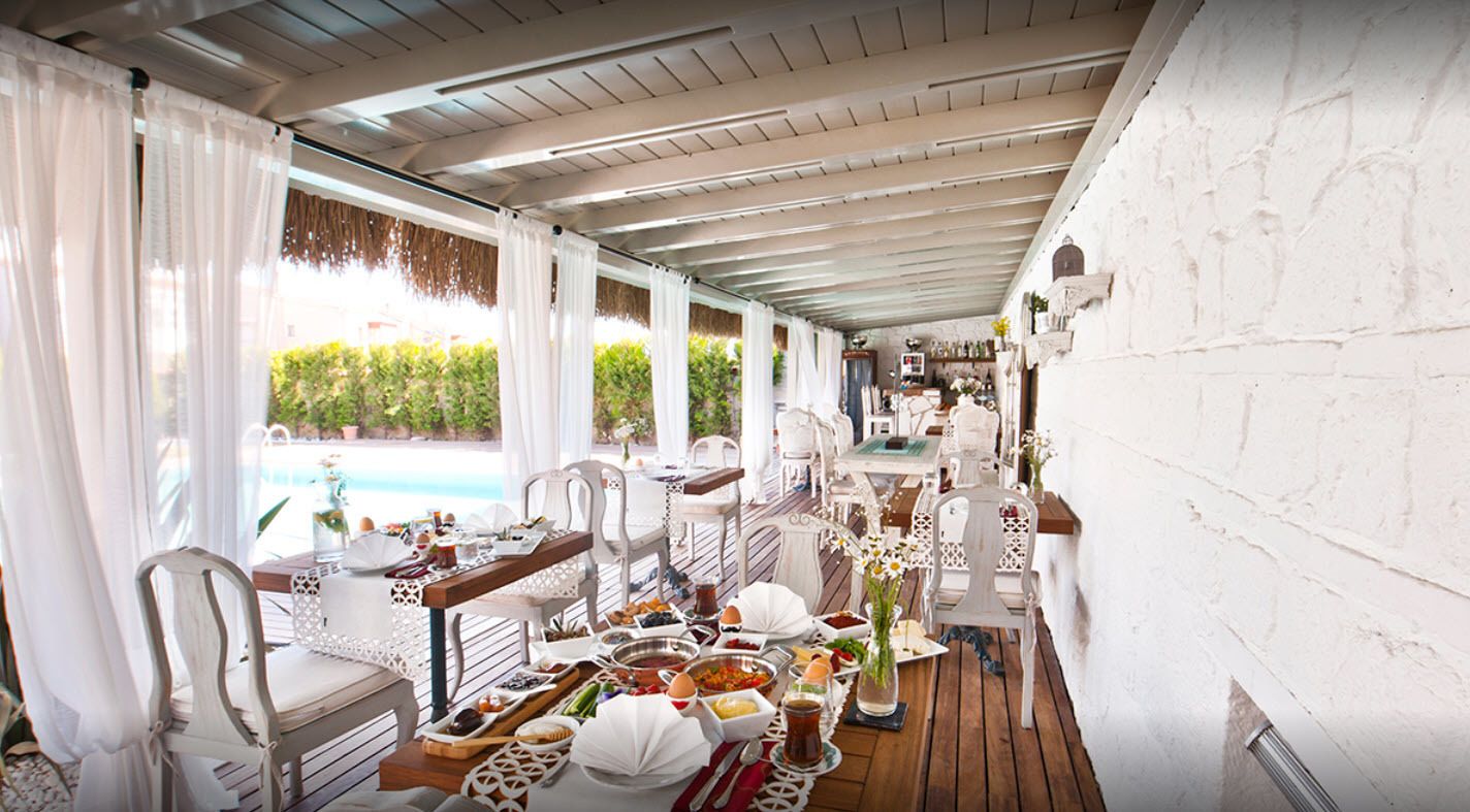 Uma área de refeições ao ar livre coberta, com mobiliário branco, vista para a piscina e decoração rústica.