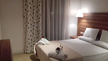 Quarto de hotel com cama bem arrumada, mesa de cabeceira com abajur e janela com cortinas.