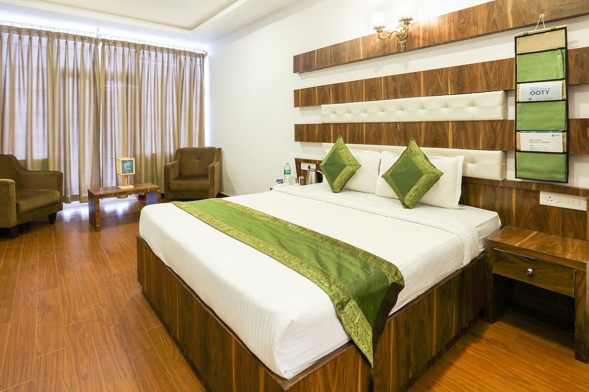 Quarto de hotel com uma cama grande, duas poltronas, uma mesa de cabeceira, uma cabeceira de madeira e uma prateleira na parede com um aviso.