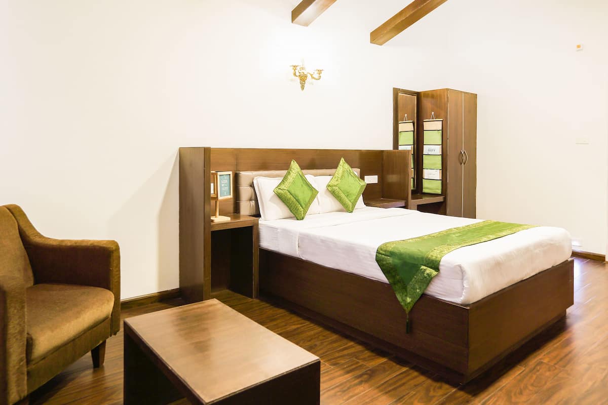 Quarto de hotel com cama, mesa de cabeceira de madeira, poltrona marrom e pequena mesa de madeira.