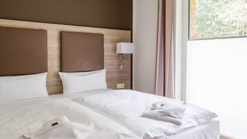 Quarto de hotel com uma cama grande, duas almofadas, uma mesa de cabeceira com abajur e uma janela com persianas.