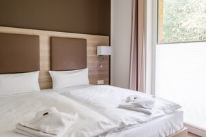 Quarto de hotel com uma cama grande, duas almofadas, uma mesa de cabeceira com abajur e uma janela com persianas.