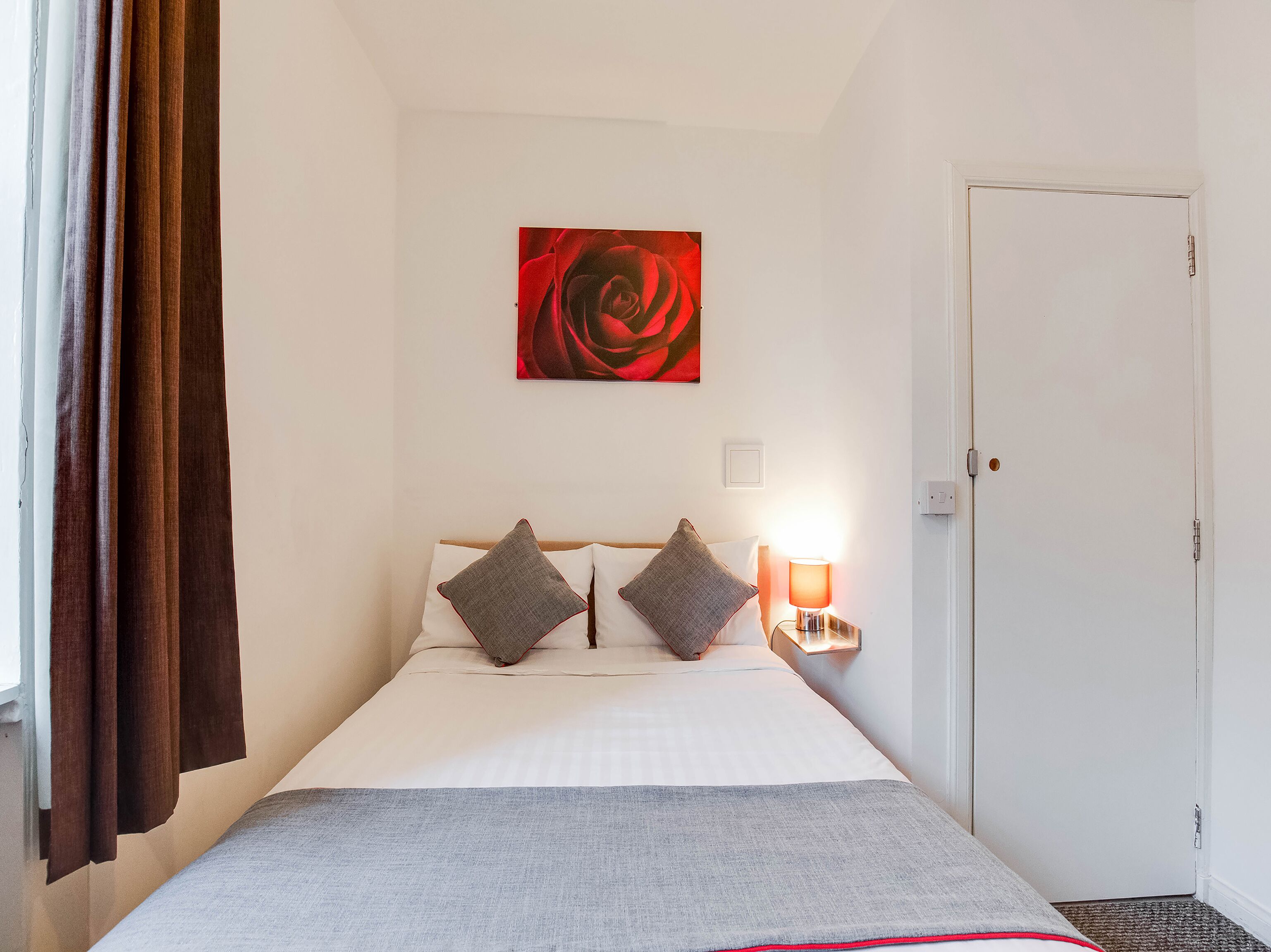 Quarto de hotel com cama, duas almofadas, abajur, porta e pintura de rosa na parede.