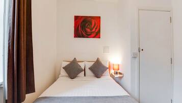 Quarto de hotel com cama, duas almofadas, abajur, porta e pintura de rosa na parede.