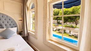 Quarto de hotel com cama, uma grande janela com vista para uma piscina e área de estar externa, e uma cabeceira decorativa.