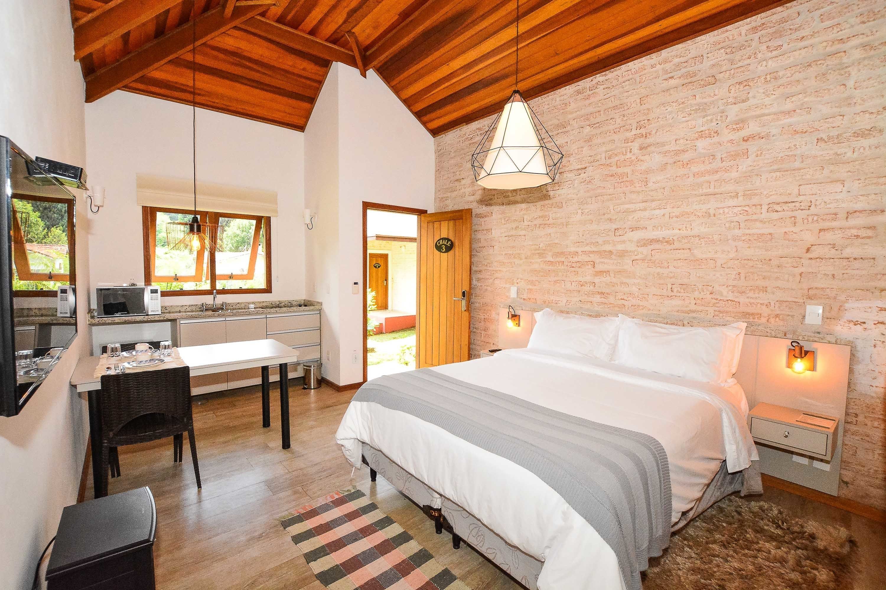 Quarto com cama grande, teto de madeira, parede de tijolos e pequena cozinha.