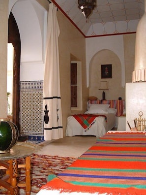 Quarto com uma cama, uma mesa e uma cadeira. Há uma janela com cortinas e uma parede com um padrão decorativo.