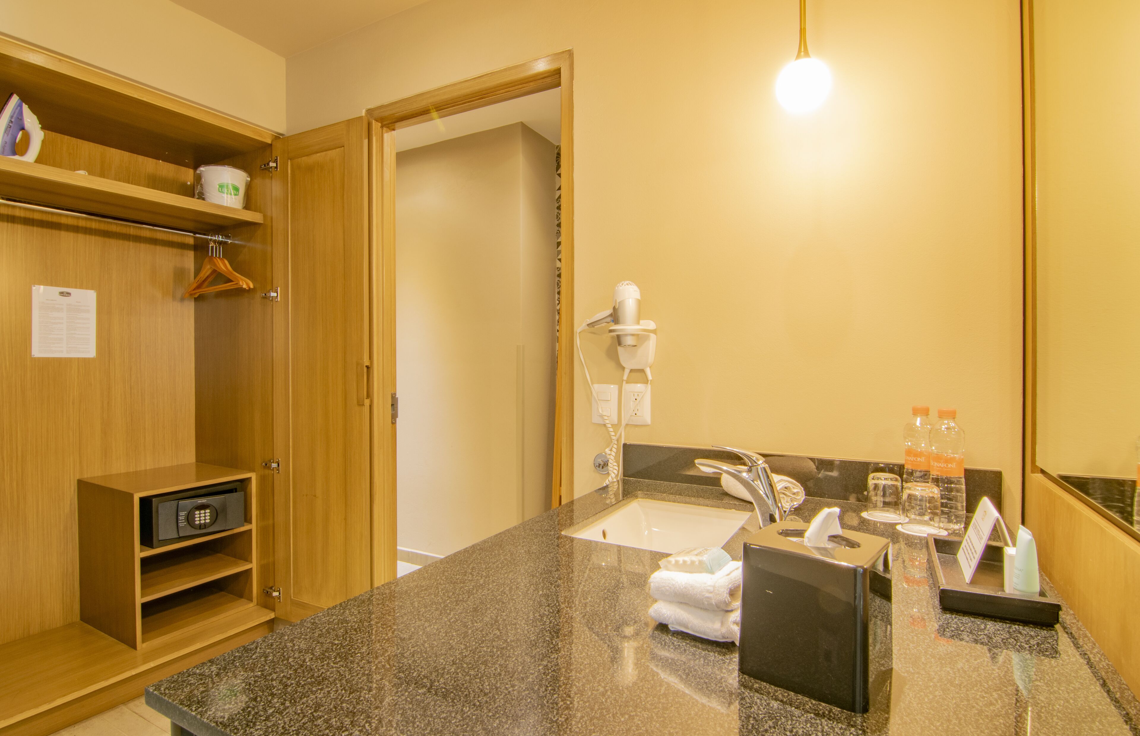 Casa de banho de hotel com bancada de granito, lavatório, espelho, porta-toalhas, secador de cabelo e um pequeno armário.