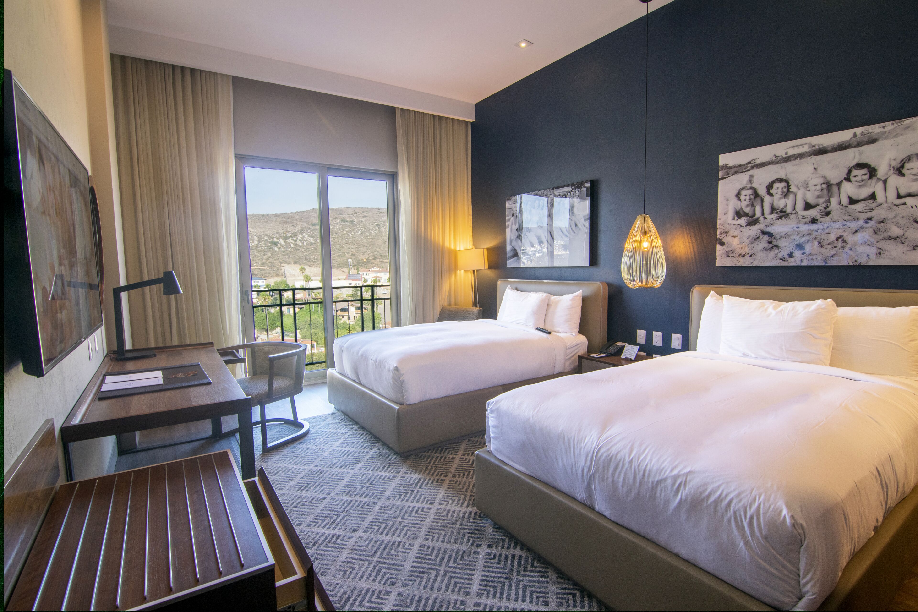 Quarto de hotel com duas camas, uma mesa, uma cadeira, uma televisão e varanda com vista para as colinas.