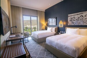 Quarto de hotel com duas camas, uma mesa, uma cadeira, uma televisão e varanda com vista para as colinas.