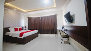 Quarto de hotel com uma cama grande, uma escrivaninha, uma cadeira, uma TV fixada na parede e uma janela com cortinas.