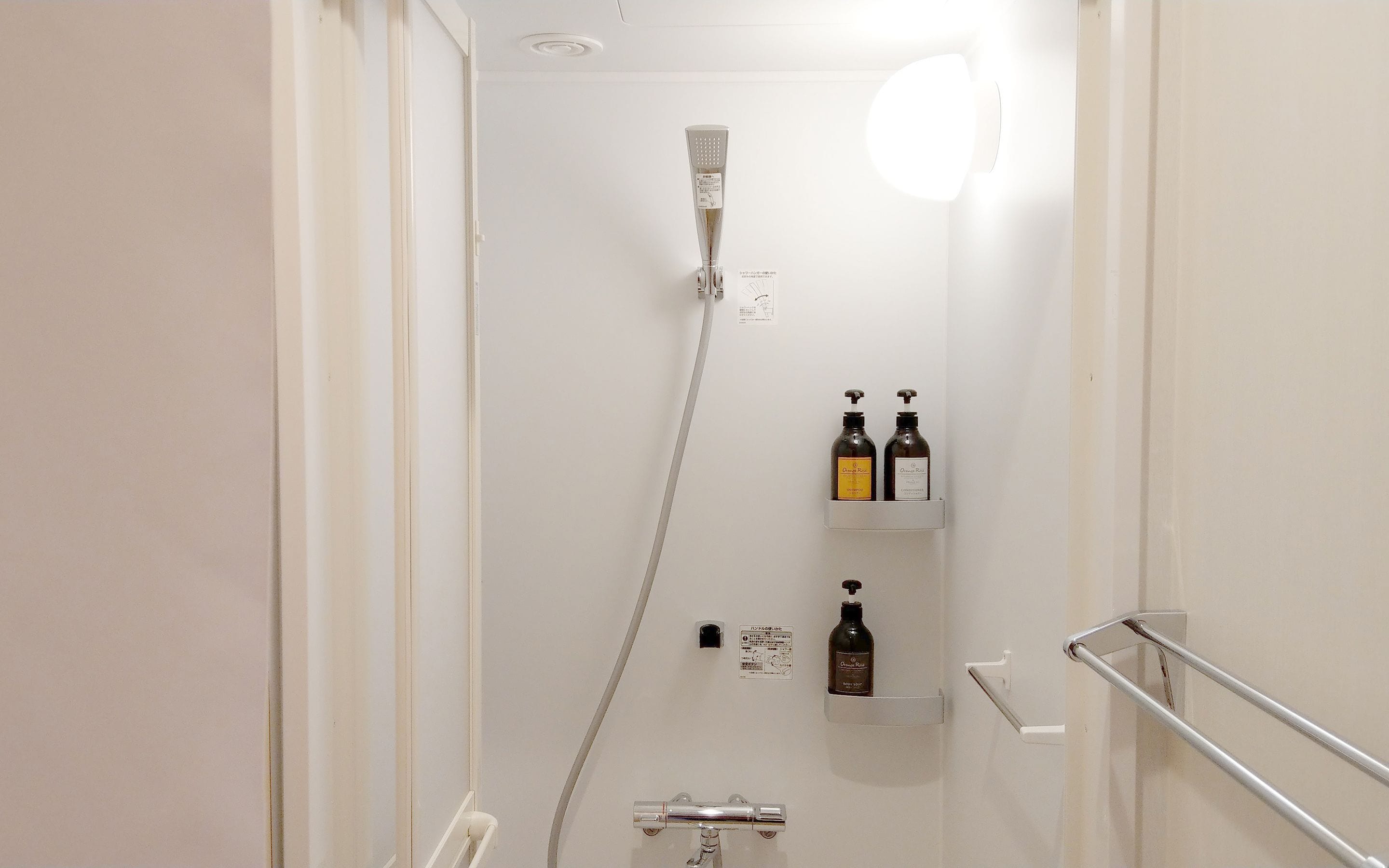 Box de chuveiro estreito com chuveiro manual, torneira fixa na parede e duas prateleiras com garrafas de shampoo e condicionador.