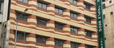 Um edifício de vários andares com fachada listrada, uma placa verde indicando 'TRIPLE BEDS' e uma entrada visível com uma placa para 'TAJIN SAFE INSURANCE'.