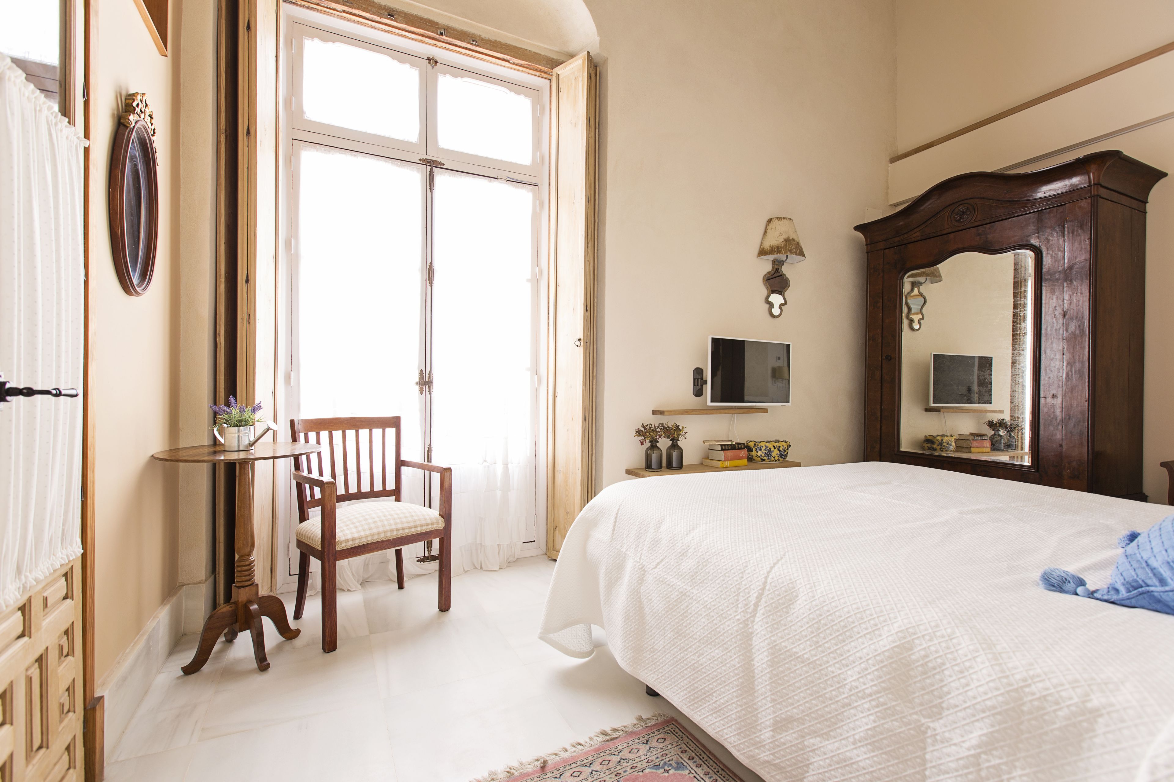 Quarto com uma cama grande, uma cadeira de madeira, uma mesa pequena com um vaso, um espelho e uma televisão em uma prateleira.