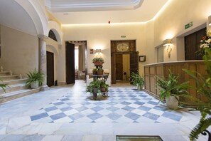 Um hall com piso xadrez, colunas, balcão de recepção e plantas em vasos.