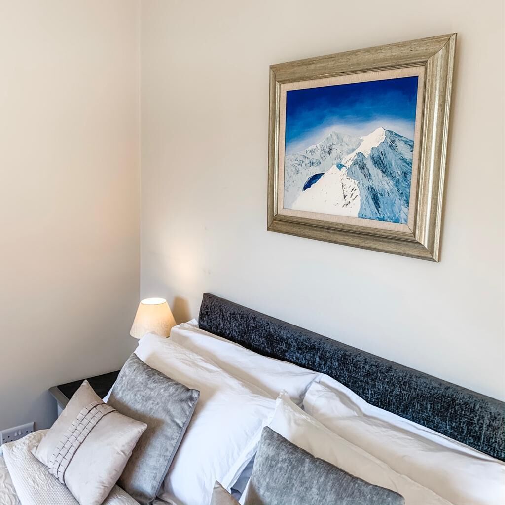 Quarto com cama, almofadas, mesa de cabeceira, abajur e um quadro com paisagem de montanhas na parede.