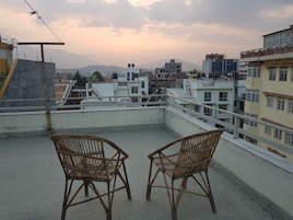 Terraço no telhado com duas cadeiras de vime, vista de um horizonte urbano com edifícios e montanhas ao fundo.