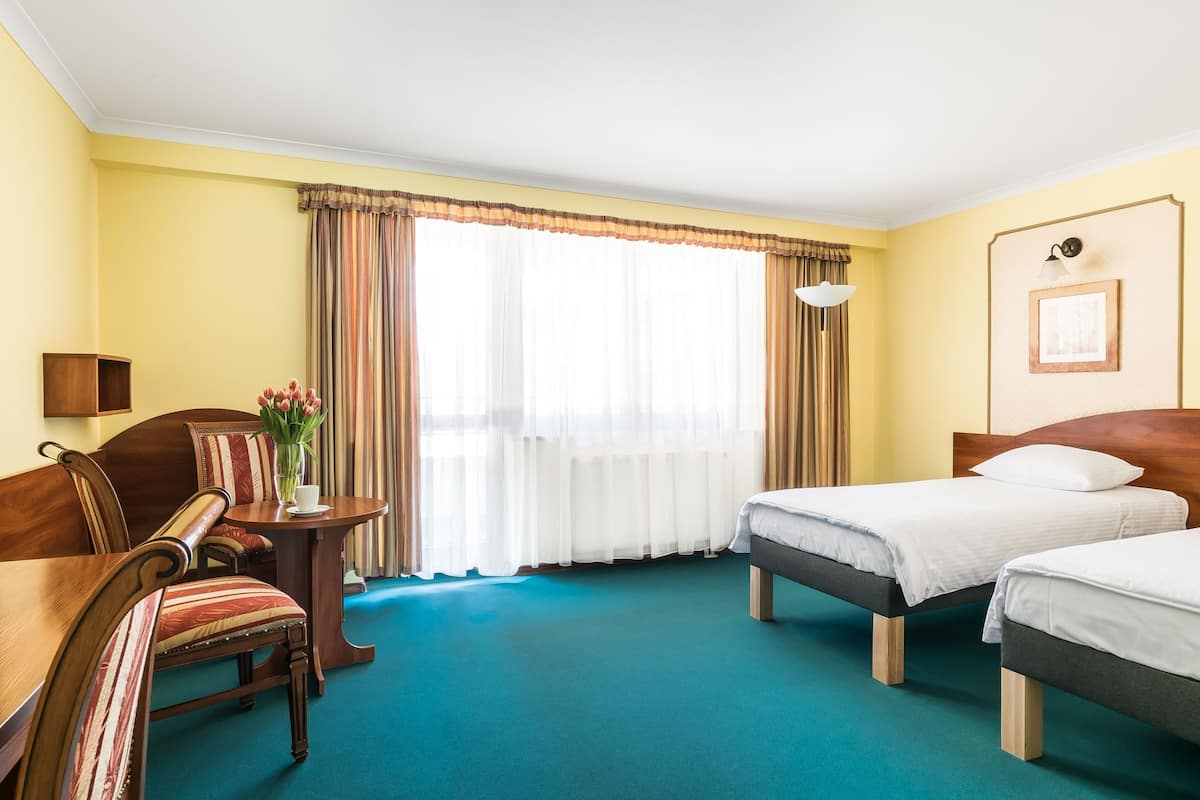 Quarto de hotel com duas camas de solteiro, uma cadeira de madeira, uma mesa pequena com um vaso de flores e uma janela grande com cortinas.