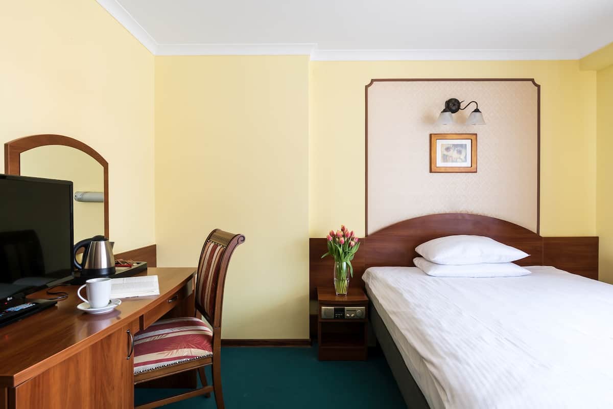 Quarto de hotel com escrivaninha de madeira, uma cadeira, cama com roupa de cama branca, mesa de cabeceira com abajur e um quadro na parede.