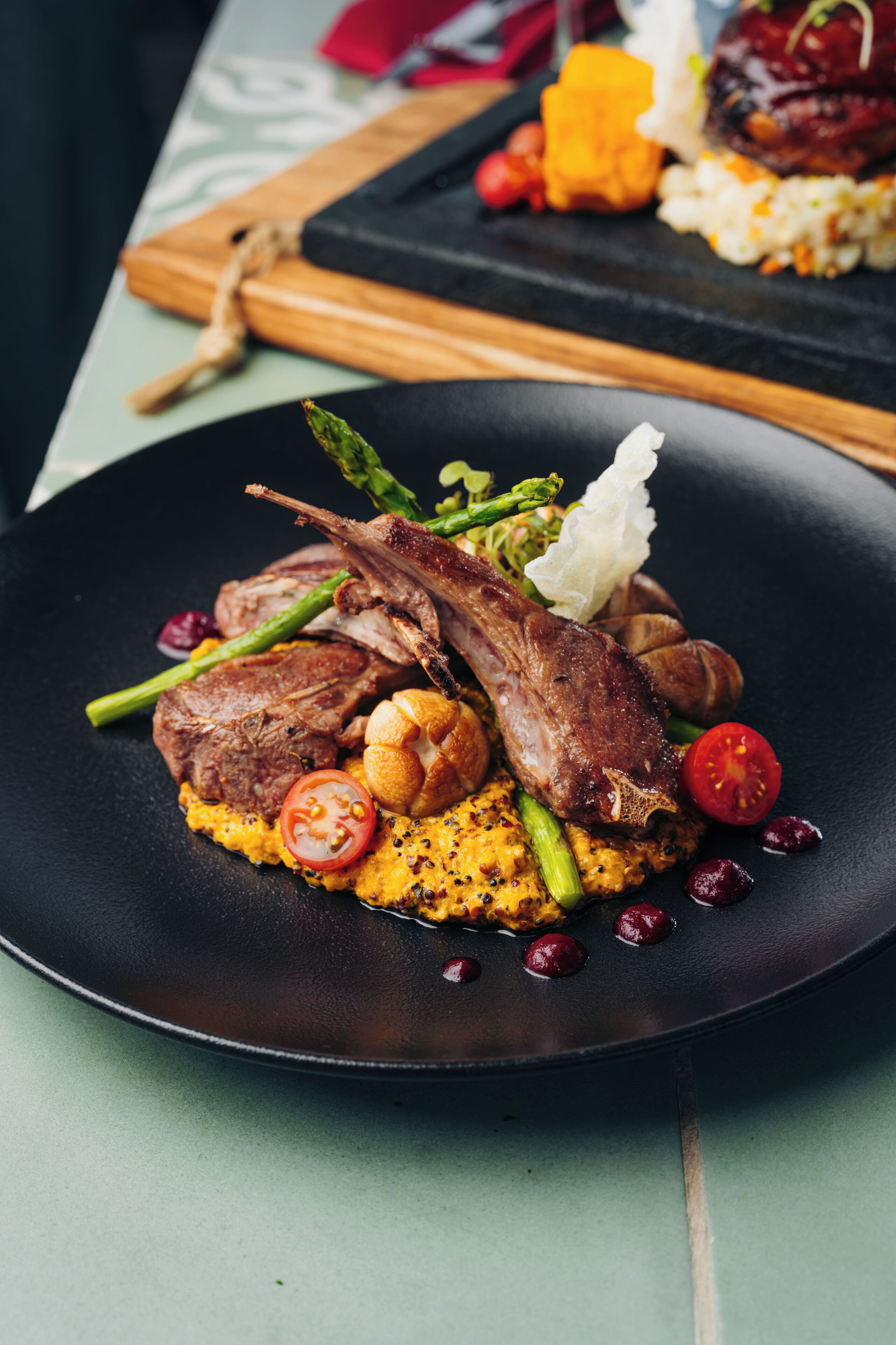Um prato com um bife de cordeiro, aspargos, tomates cereja e molho amarelo.