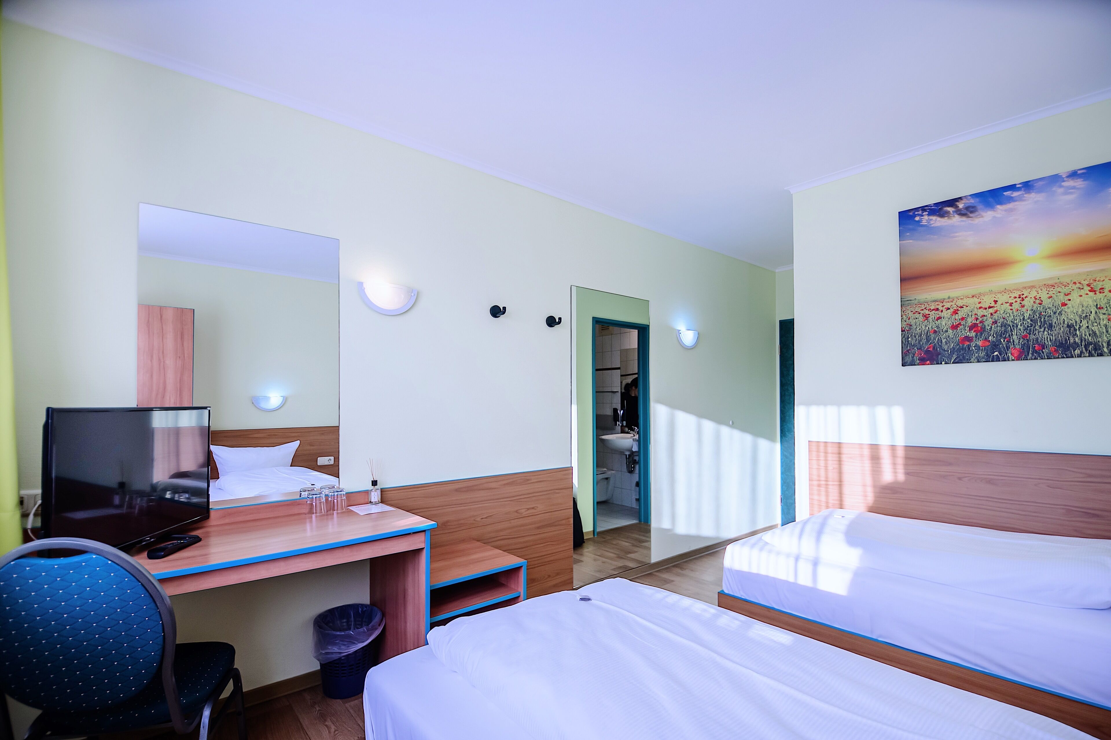 Quarto de hotel com cama, escrivaninha com computador, cadeira e banheiro visível por uma porta aberta.